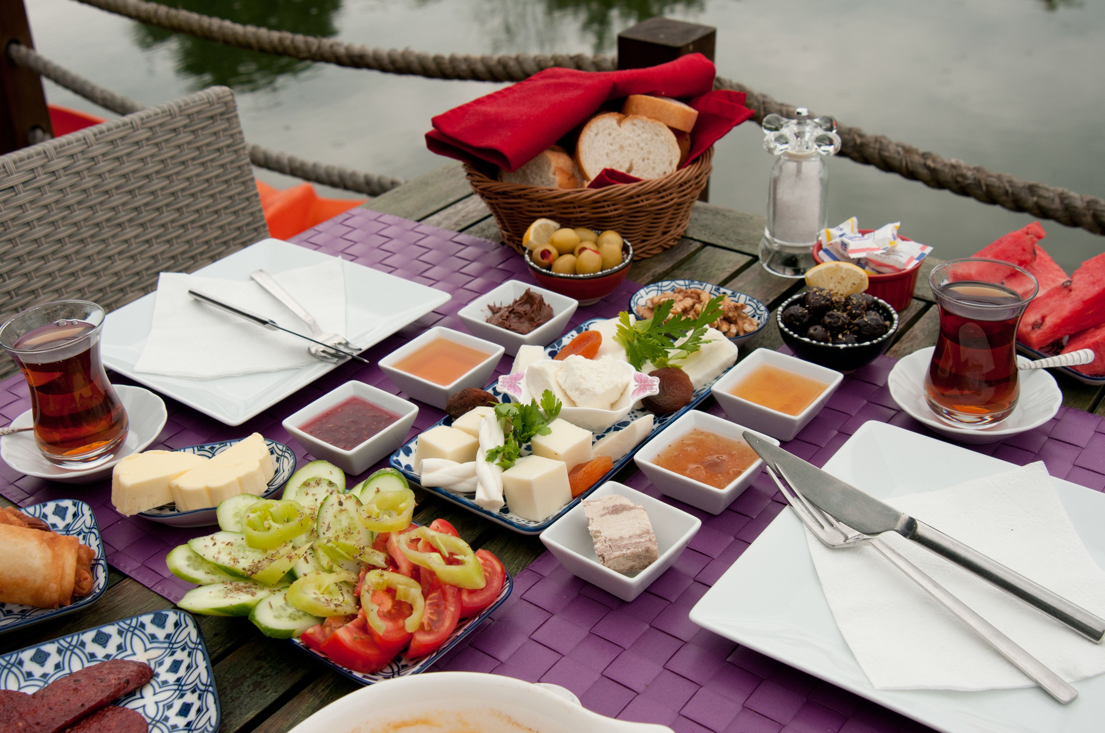 Mesa posta com diversos alimentos e bebidas, incluindo queijo, azeitonas e pão, em um deck de madeira à beira de um corpo d'água.