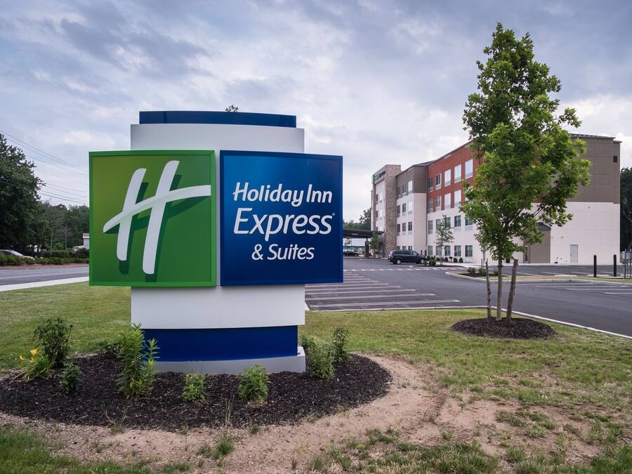 Placa do Holiday Inn Express & Suites com esquema de cores azul e verde, cercada por uma área ajardinada com plantas pequenas e uma árvore.