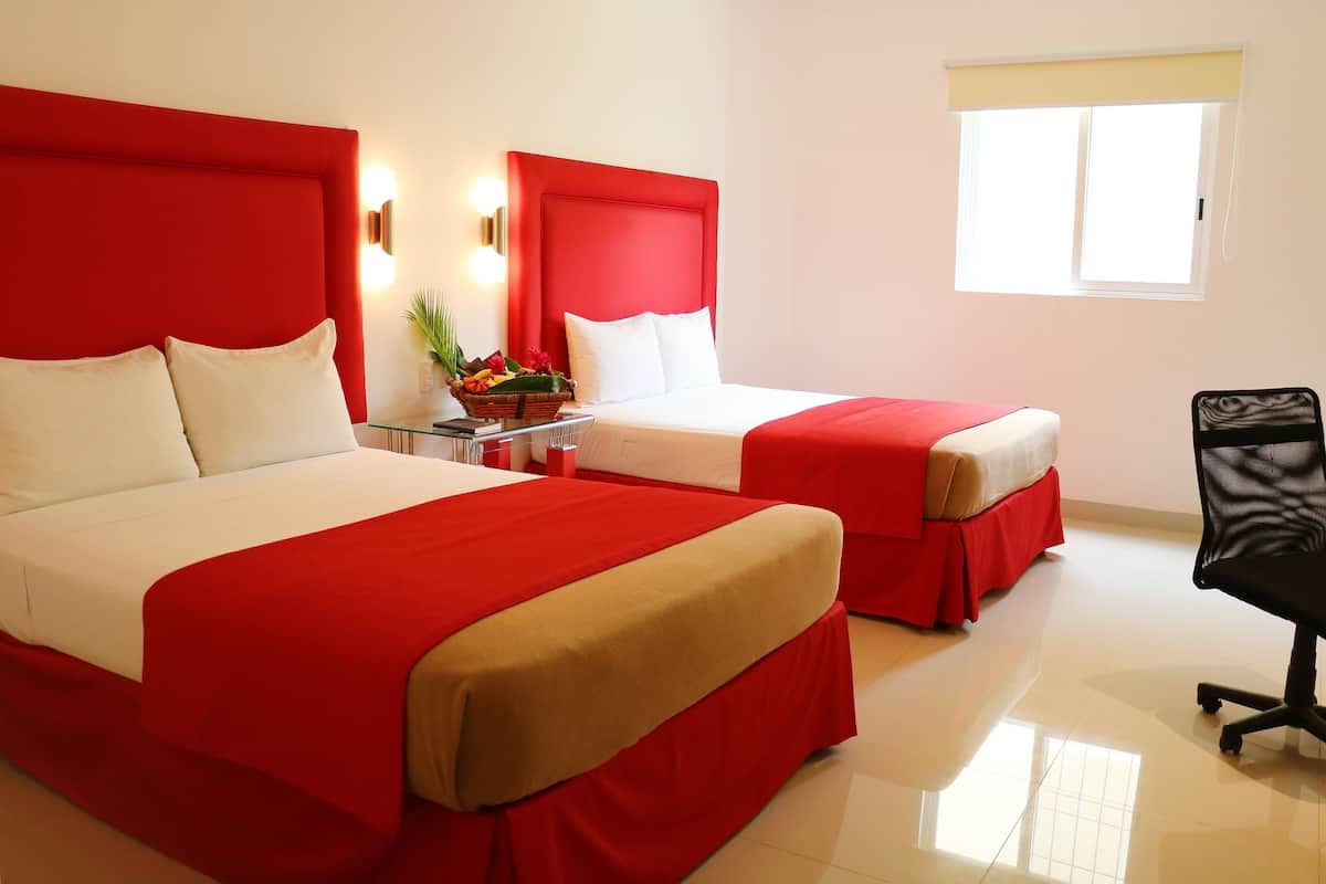 Quarto de hotel com duas camas, uma mesa pequena com cesta de frutas, uma cadeira e uma janela com persianas.