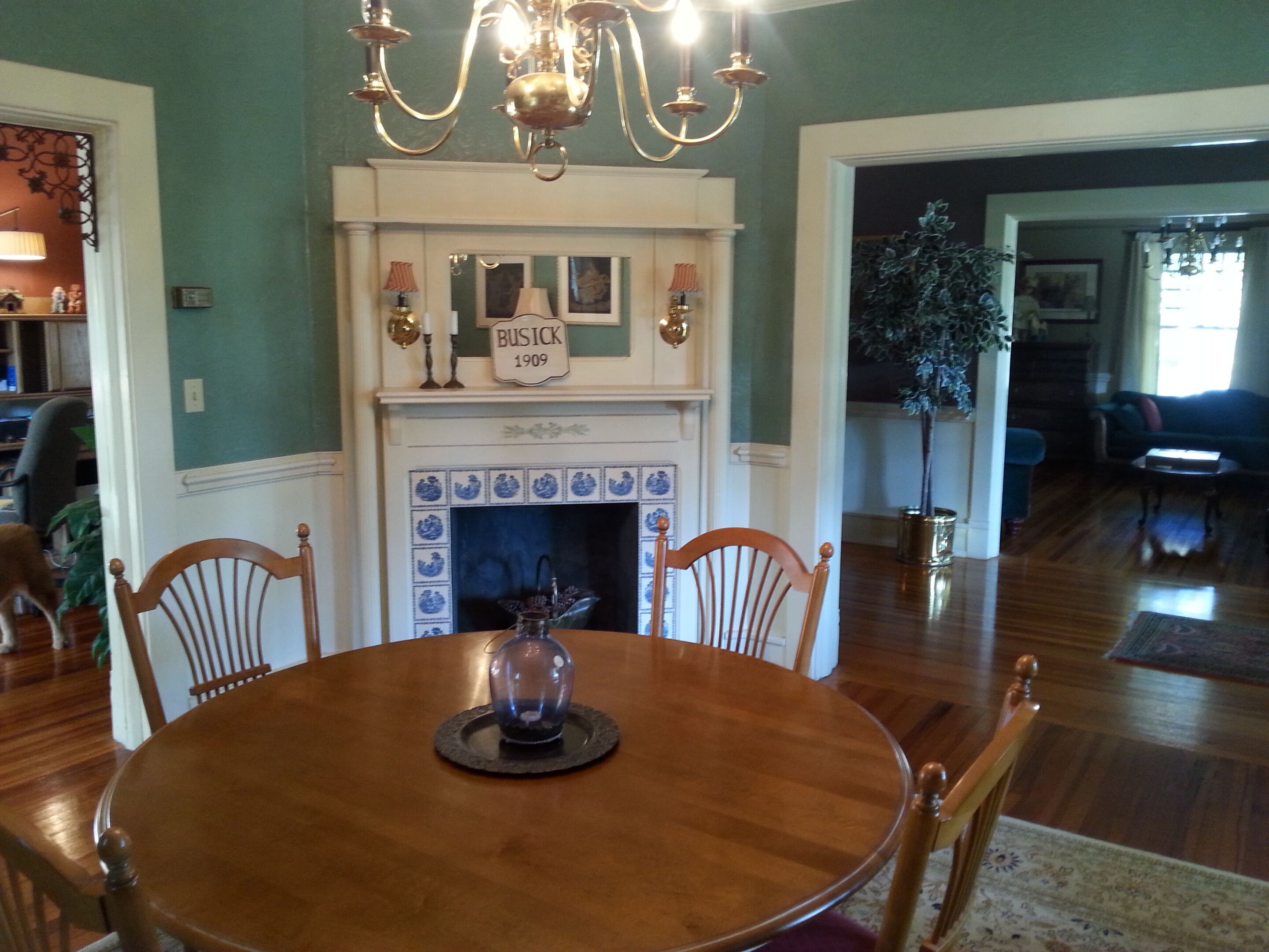 Uma sala de jantar com uma mesa redonda de madeira, cadeiras, um lustre, uma lareira com o letreiro 'BUSICK 1909' e um vaso de plantas.