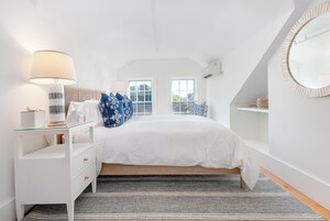 Um quarto com cama, mesa de cabeceira, abajur, espelho e janela.