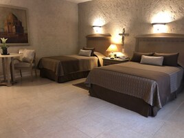 Quarto de hotel com duas camas, uma escrivaninha e uma cadeira. Há luminárias na parede e um quadro pendurado.