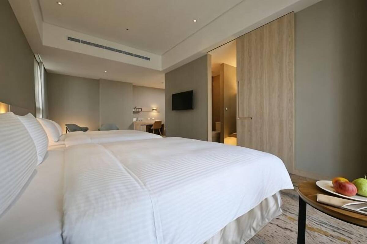 Quarto de hotel com uma cama grande, uma escrivaninha com cadeira, televisão e um prato de frutas no criado-mudo.