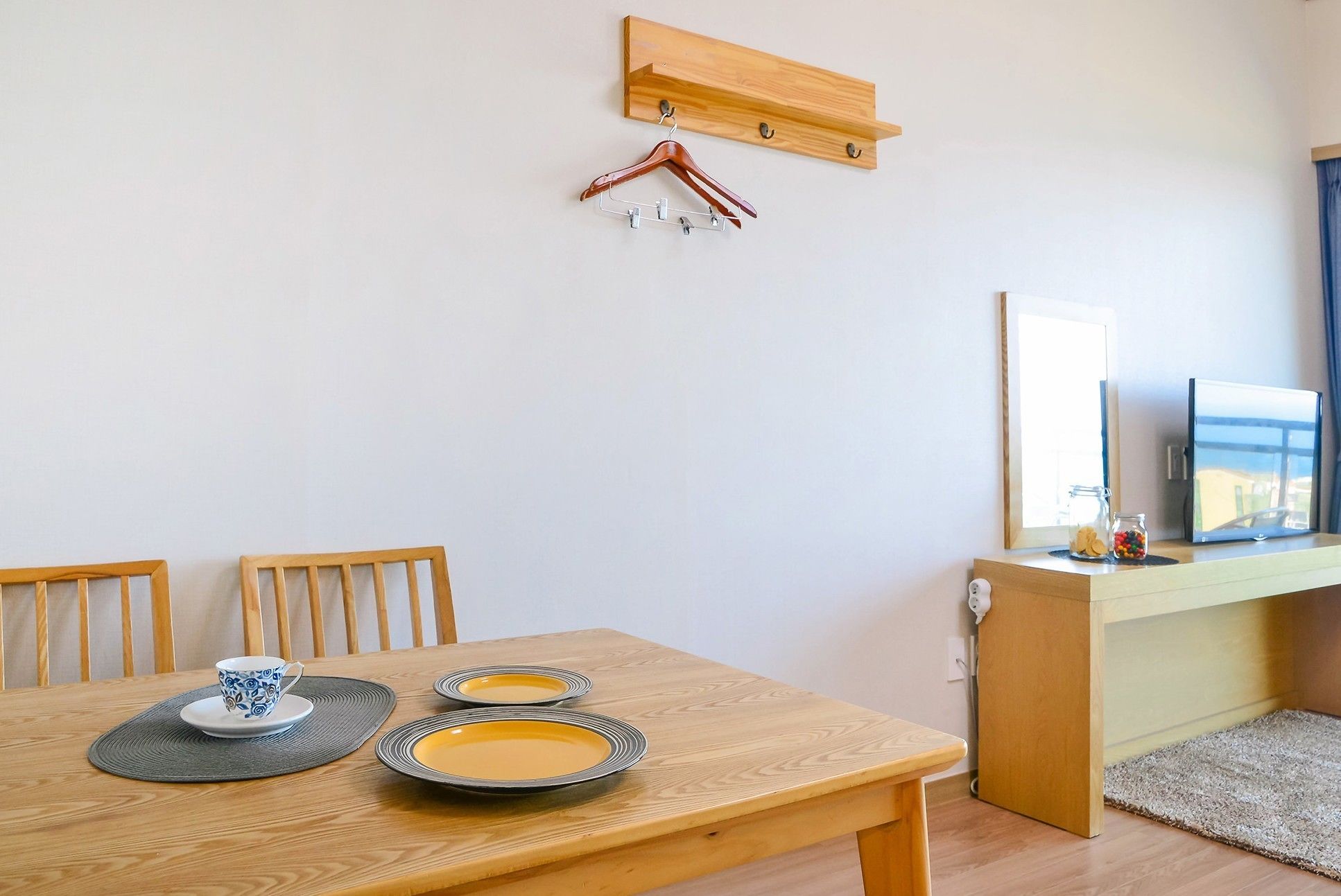 Uma área de jantar com mesa de madeira posta para dois, um cabideiro de madeira acima e uma pequena cozinha com espelho e televisão.