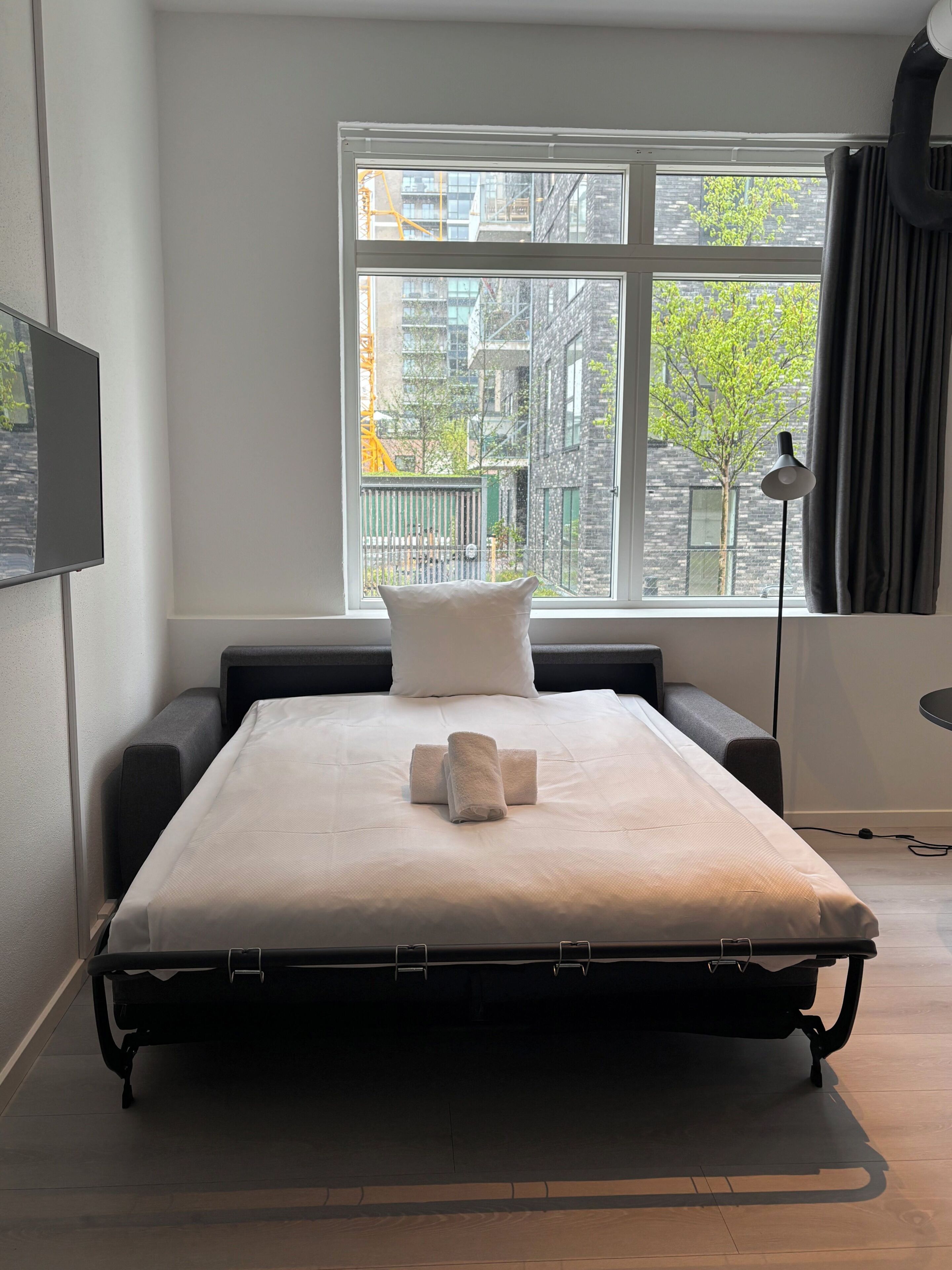 Quarto de hotel moderno com cama, TV, janela com vista para edifícios e árvores, e um abajur.