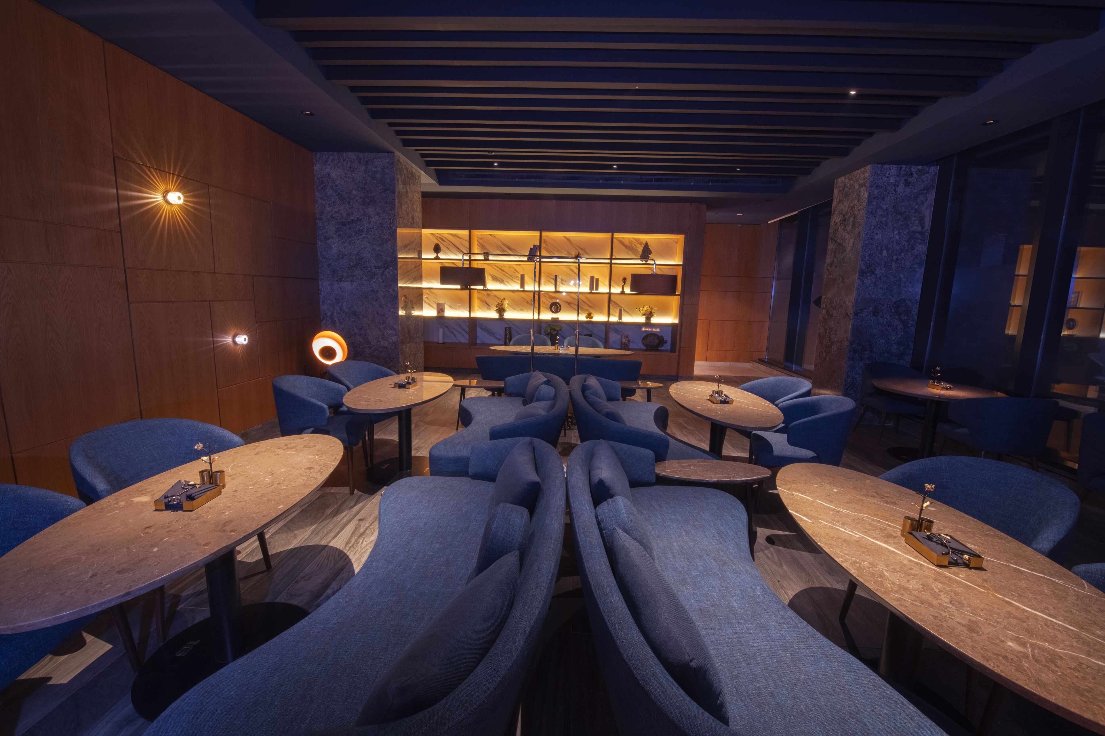 Espaço lounge moderno com assentos estofados em azul e mesas redondas, com uma unidade de estantes central decorativa.