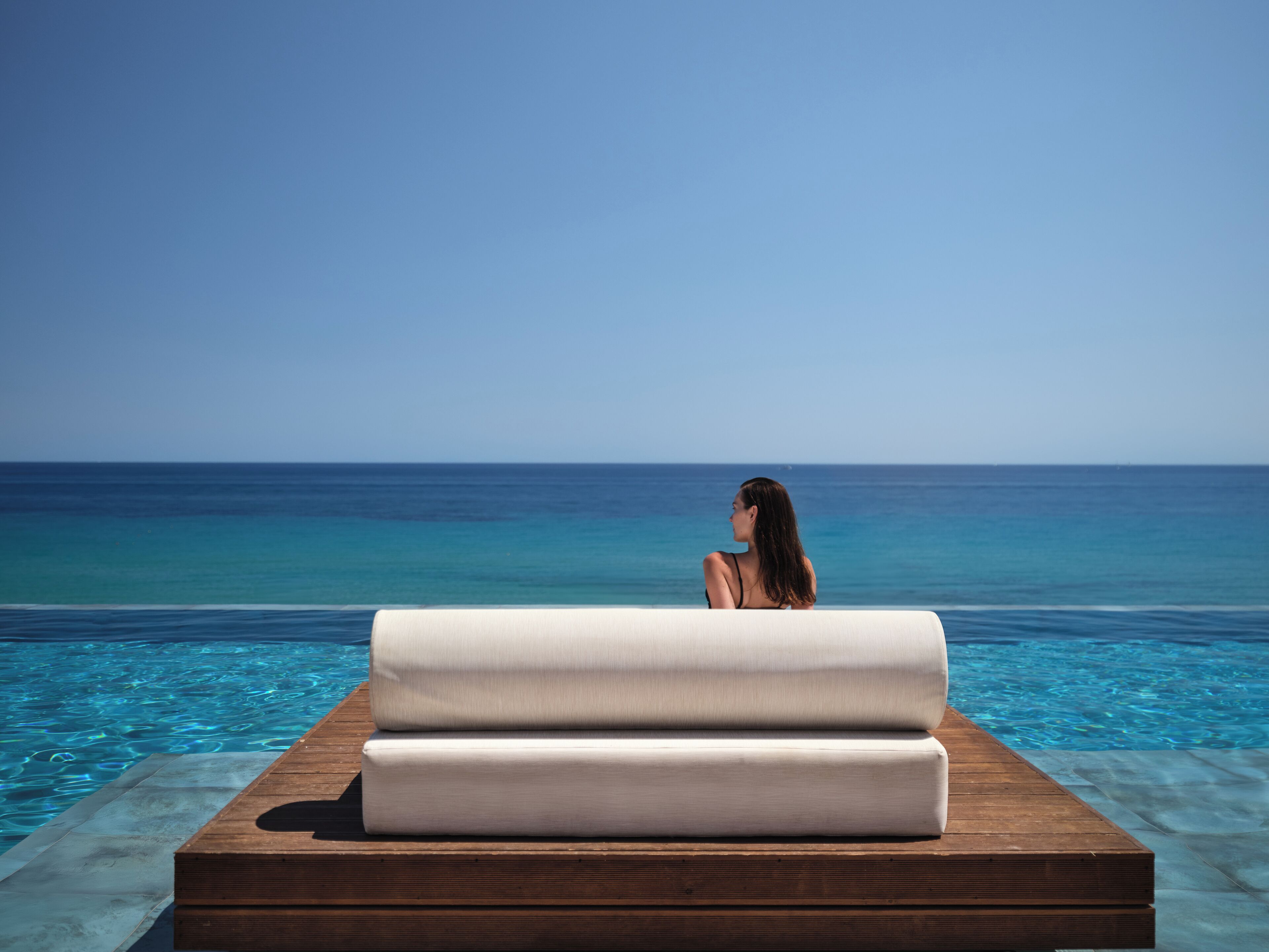 Um banco de madeira com almofada branca à beira da piscina, com vista para o oceano azul e límpido.