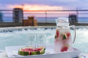 Jarra de vidro com fatias de melancia e um copo de água em um tabuleiro, perto de uma piscina com vista para o pôr do sol.