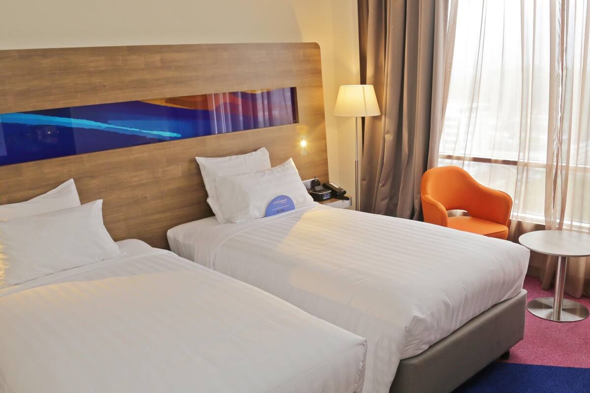 Quarto de hotel com duas camas, uma mesa de cabeceira com abajur, uma poltrona laranja e uma pequena mesa.