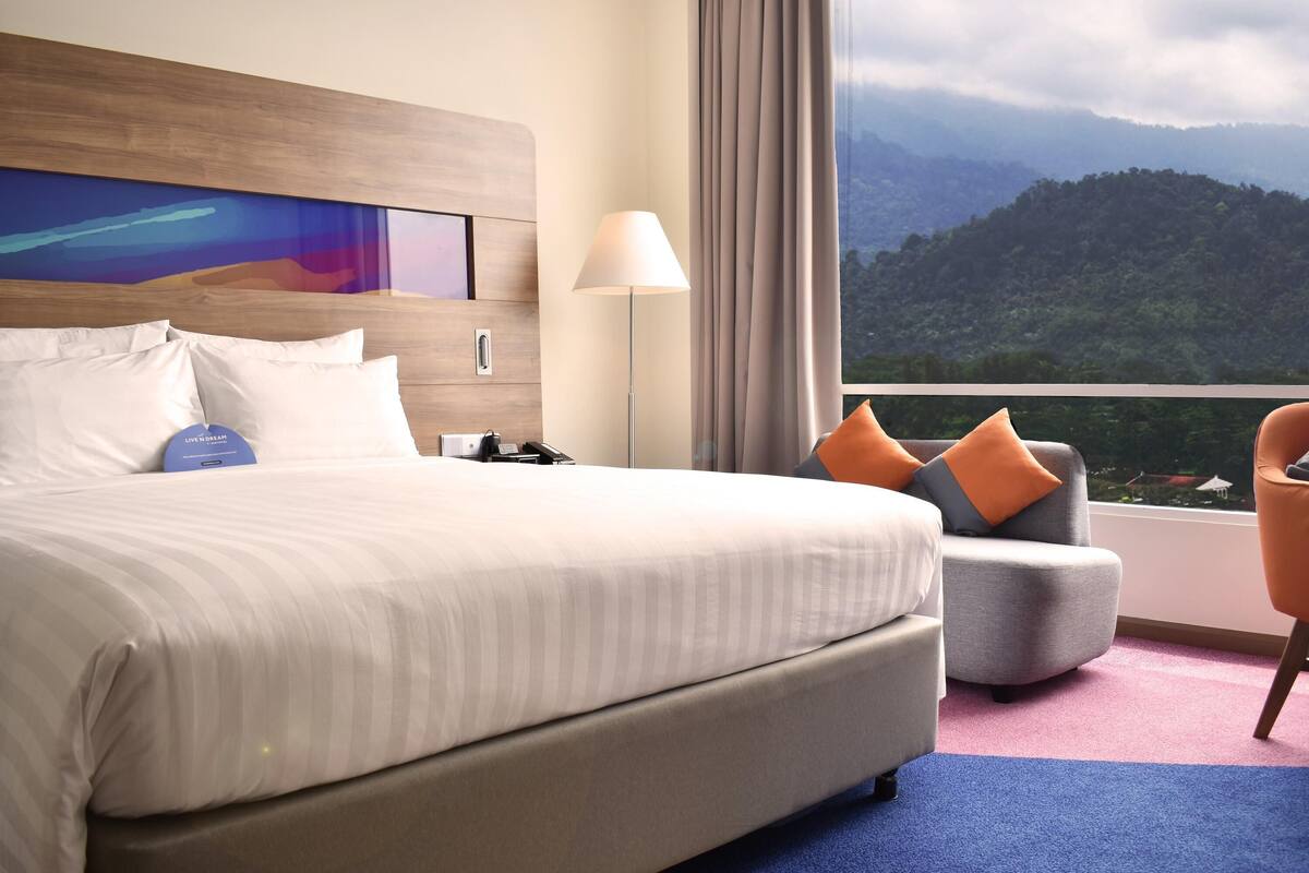 Quarto de hotel com uma cama grande, uma escrivaninha com abajur, uma cadeira e vista para uma paisagem montanhosa.