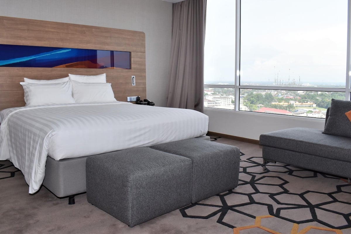 Quarto de hotel com uma cama grande, duas poltronas cinzentas, um sofá, vista para a cidade e uma pintura abstrata moderna na parede.