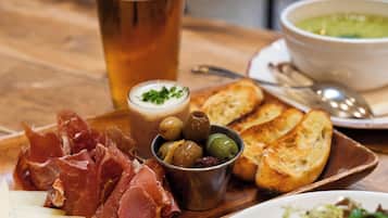 Uma mesa de madeira com uma cerveja num copo, uma tigela de sopa, presunto, azeitonas e pão tostado.