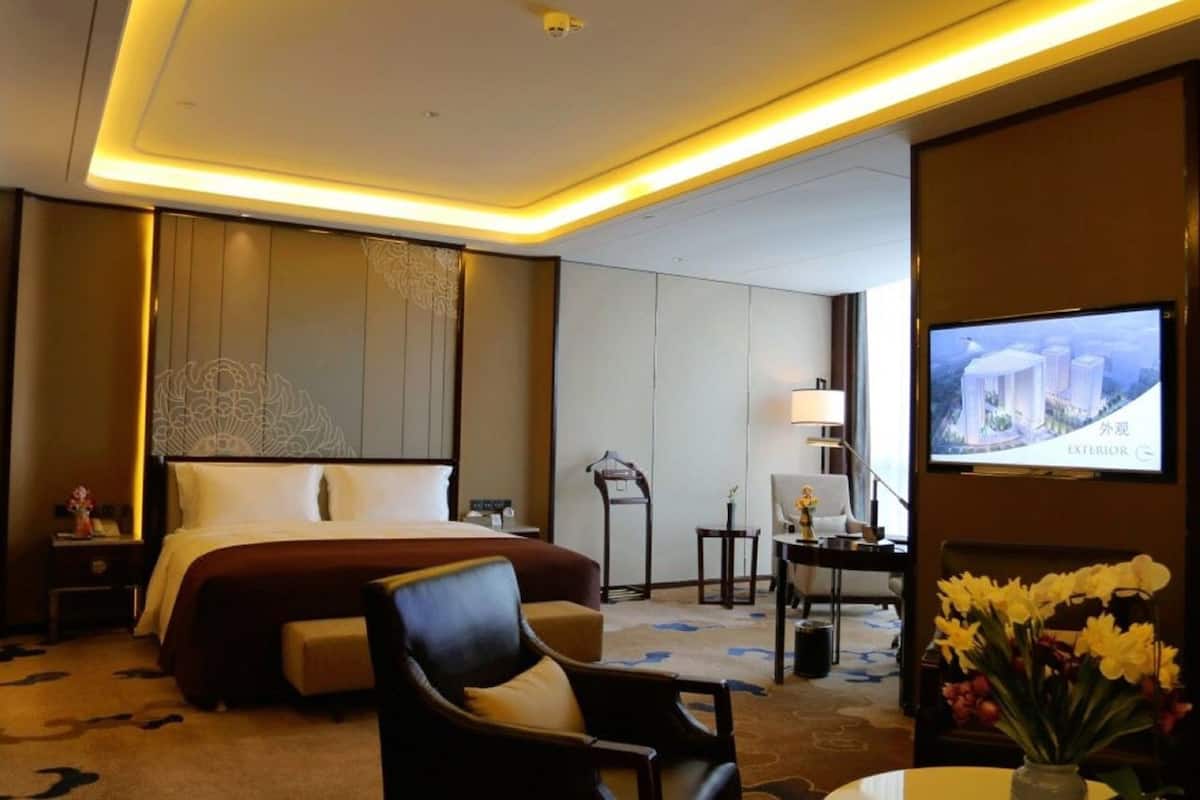 Quarto de hotel com cama grande, escrivaninha, cadeira, televisão e um vaso com flores.