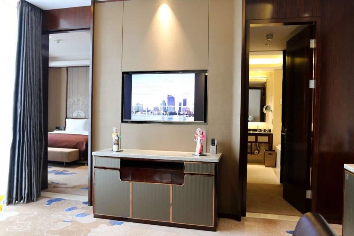Quarto de hotel com uma cama, televisão na parede, uma pequena mesa com um vaso e vista para o horizonte da cidade.