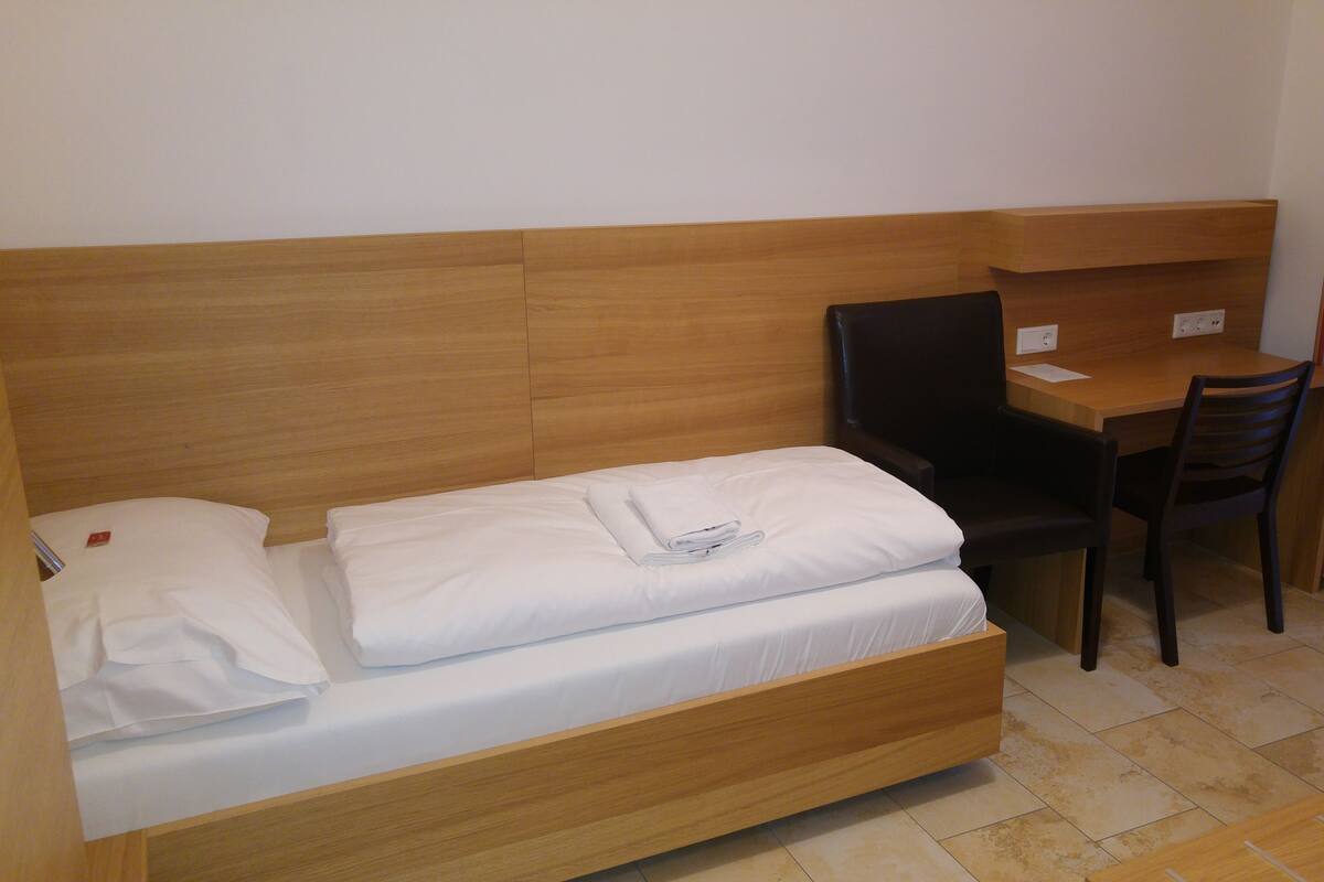 Uma cama compacta com cabeceira de madeira, uma pequena escrivaninha com cadeira e uma cortina listrada.