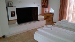 Quarto de hotel com televisão de tela plana em uma unidade de entretenimento branca, cômoda de madeira, cama com lençóis brancos e uma pintura grande na parede.