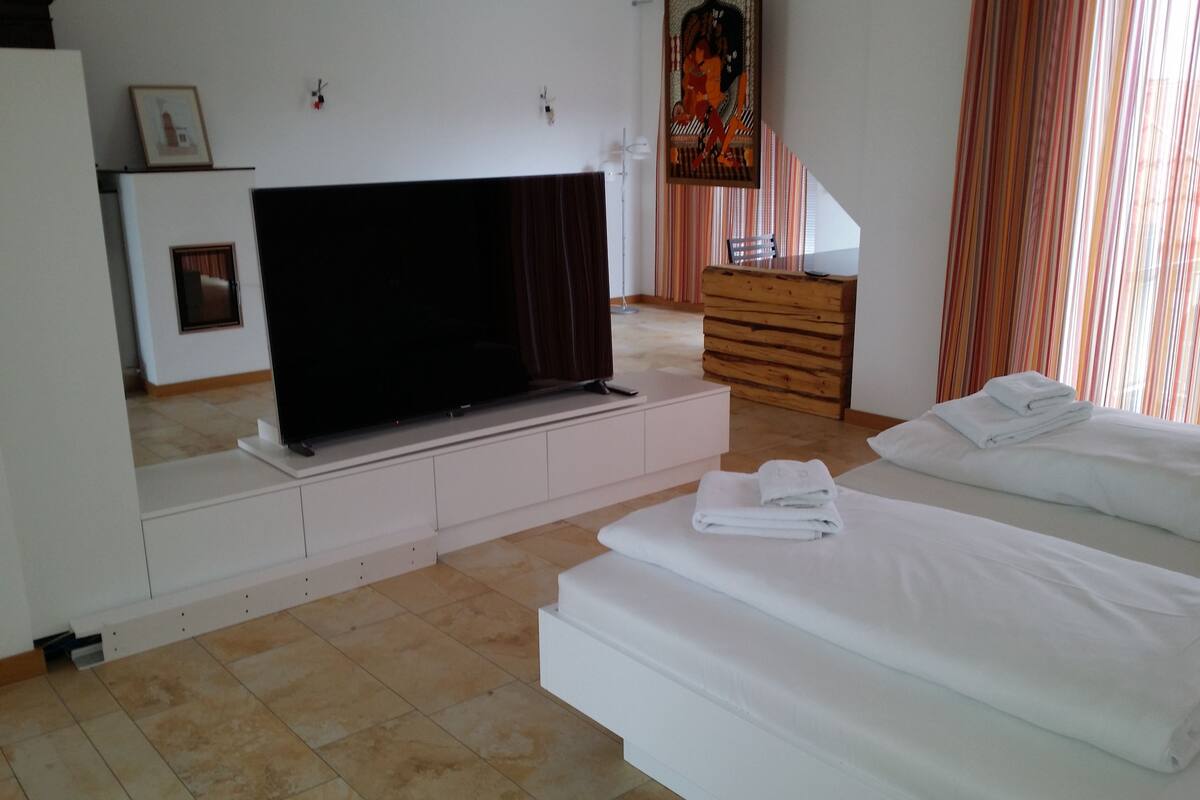 Quarto de hotel com televisão de tela plana em uma unidade de entretenimento branca, cômoda de madeira, cama com lençóis brancos e uma pintura grande na parede.