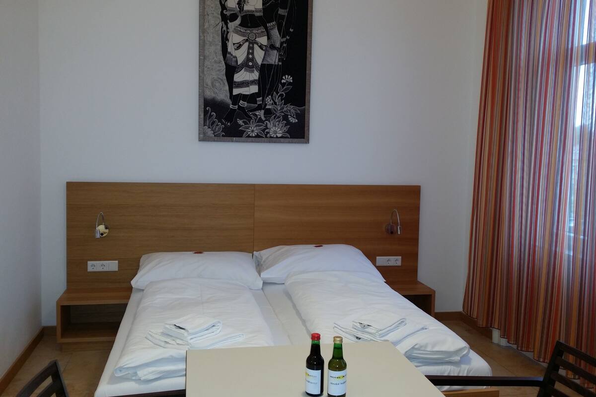 Uma cama de casal com lençóis brancos, duas garrafas de vinho na mesa de cabeceira e uma cabeceira de madeira.