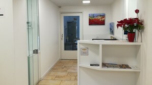Uma área de recepção com porta de vidro, balcão de recepção, um vaso de flores vermelho e um quadro na parede.