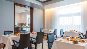 Uma sala de jantar de hotel com várias mesas preparadas para o pequeno-almoço, com sumo de laranja e diversas opções de refeições matinais.