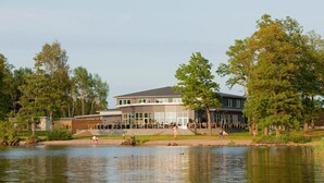 Um edifício moderno com fachada de vidro extensa, localizado ao lado de um lago tranquilo onde pessoas desfrutam atividades ao ar livre.