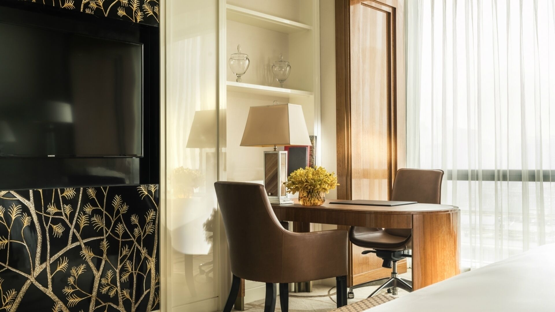 Quarto de hotel com televisão, secretária com duas cadeiras, abajur e um vaso com flores.