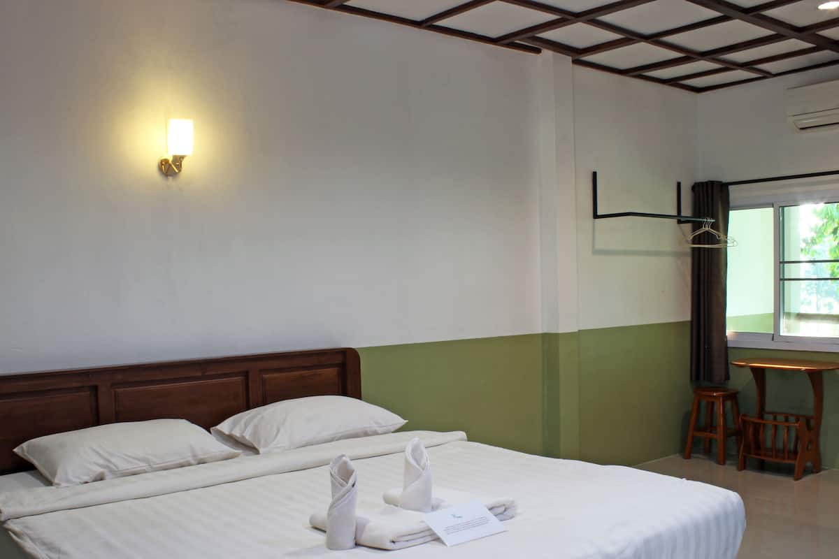 Cama bem arrumada com lençóis e toalhas brancos, cabeceira de madeira, luminária fixa na parede e janela com vista para o verde.
