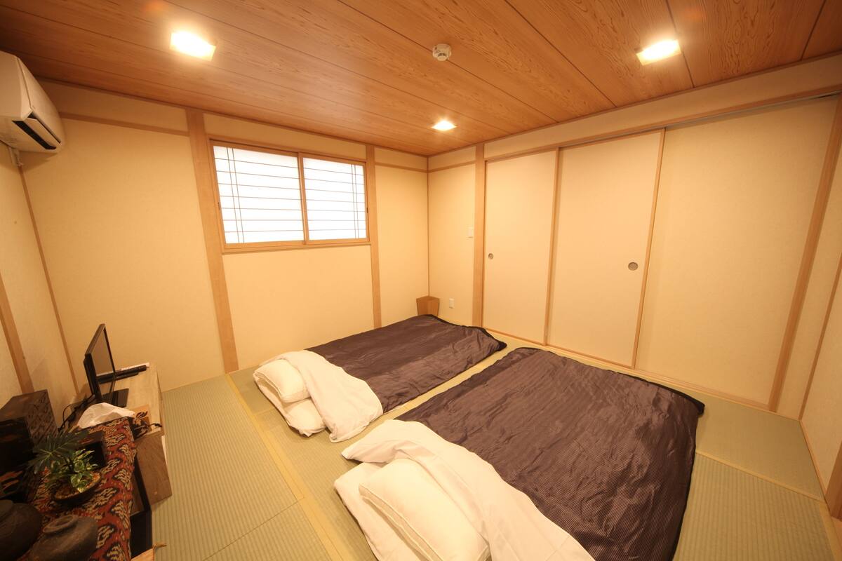 Quarto tradicional japonês com tatames, duas camas de solteiro, um futon, uma pequena mesa e um aparelho de ar condicionado.