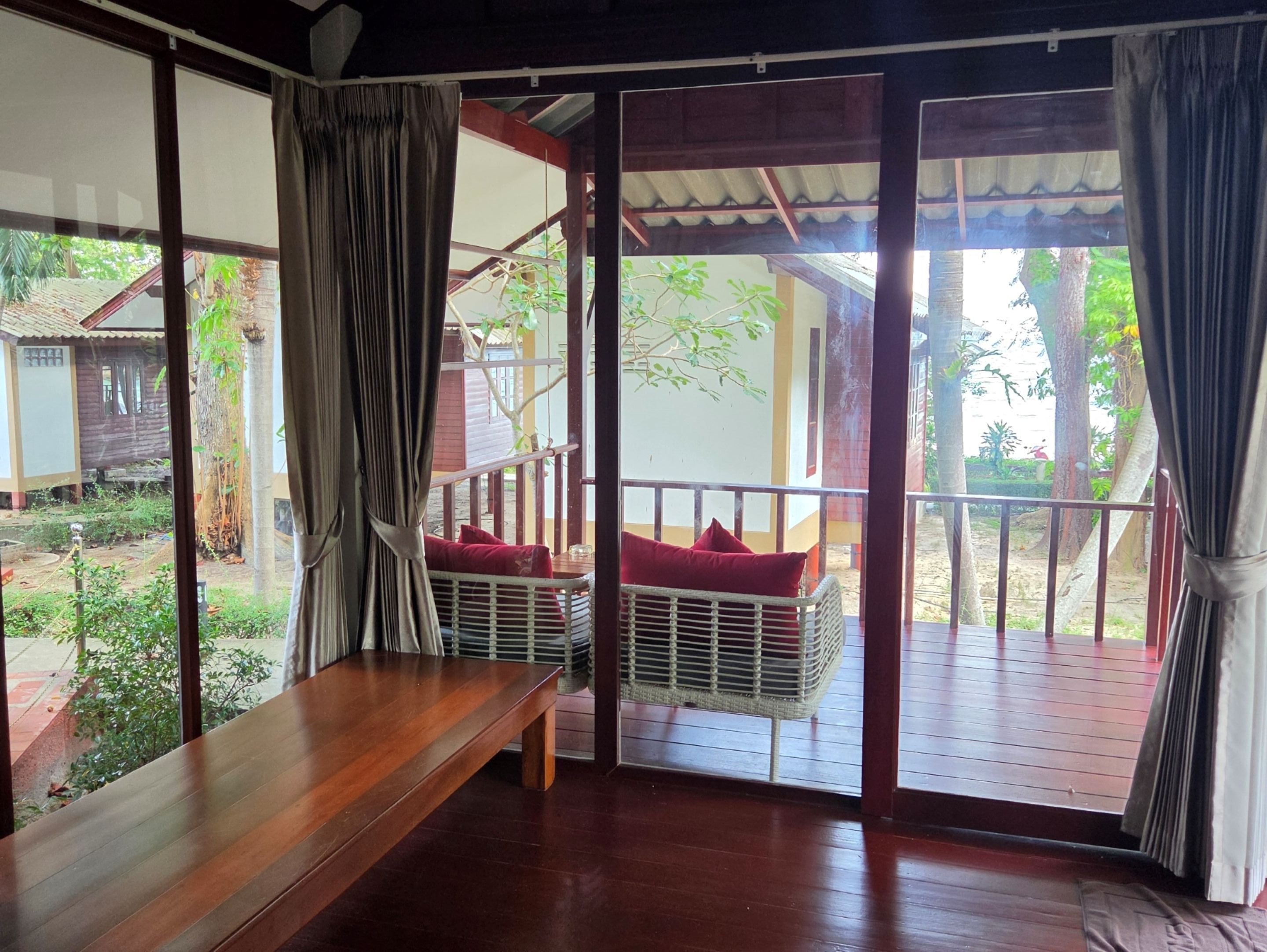 Quarto com um banco de madeira, uma cadeira de vime com almofadas vermelhas e vista para um deck de madeira e vegetação.