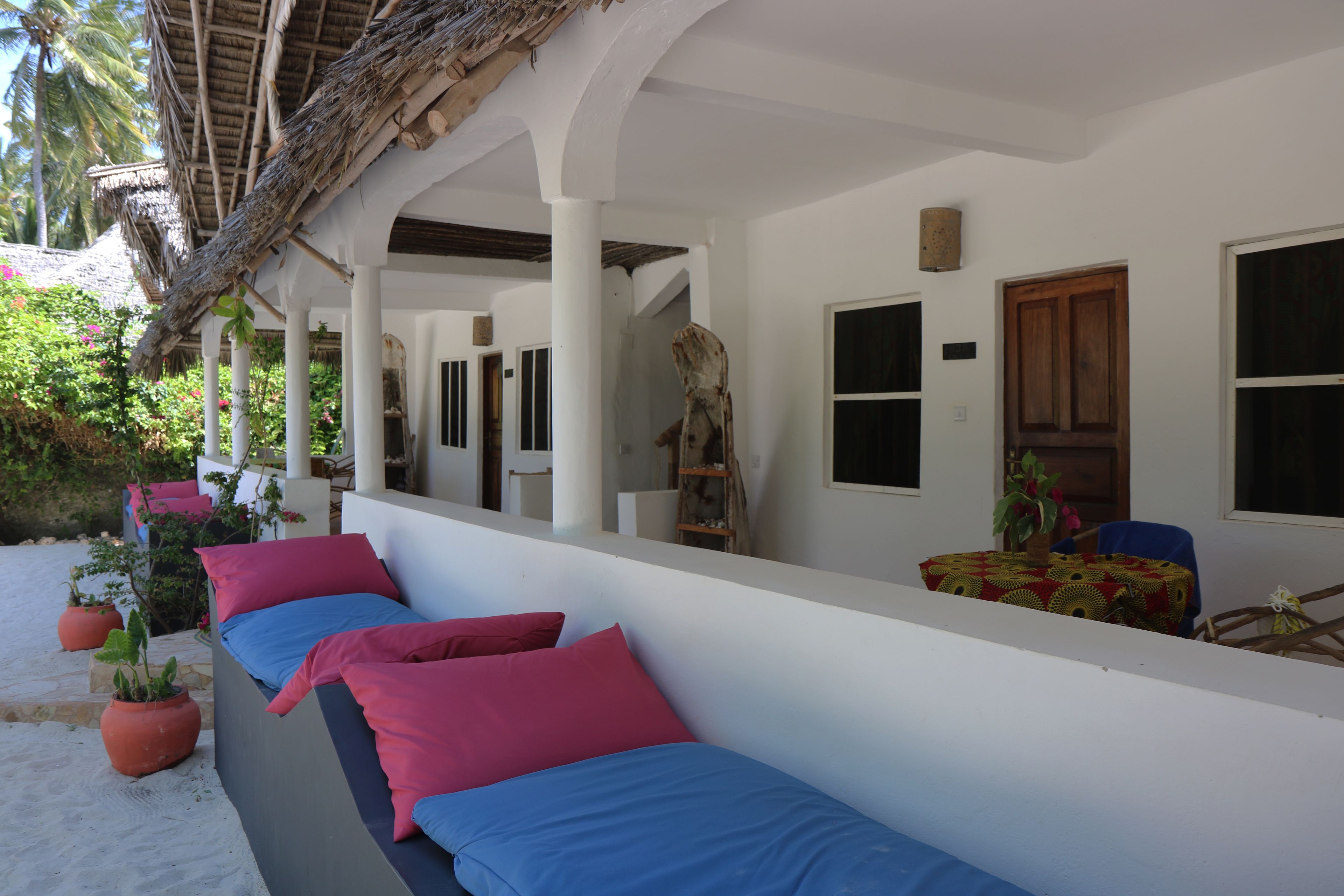 Quarto de hotel à beira-mar com telhado de palha, paredes brancas e varanda com almofadas azuis e vermelhas.