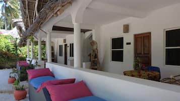 Quarto de hotel à beira-mar com telhado de palha, paredes brancas e varanda com almofadas azuis e vermelhas.