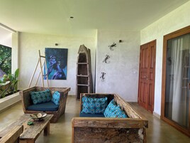 Quarto com duas sofás de madeira com almofadas estampadas, uma mesa de madeira, uma escada, um quadro na parede e uma porta com cortina.