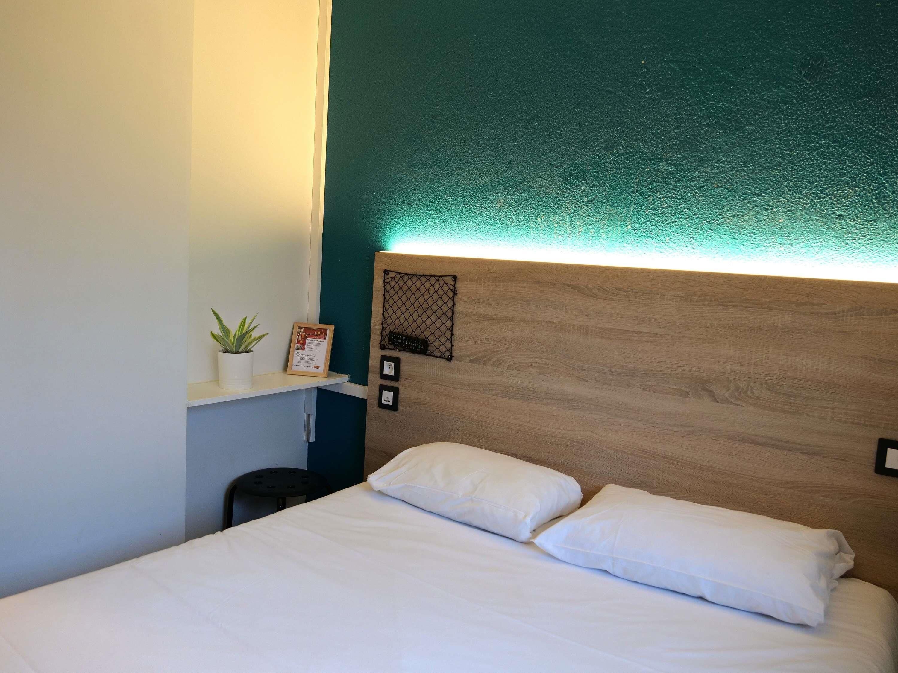 Quarto de hotel com uma cama, uma pequena prateleira com uma planta e um certificado emoldurado, e uma cabeceira com parede verde-água texturizada e painel de madeira.