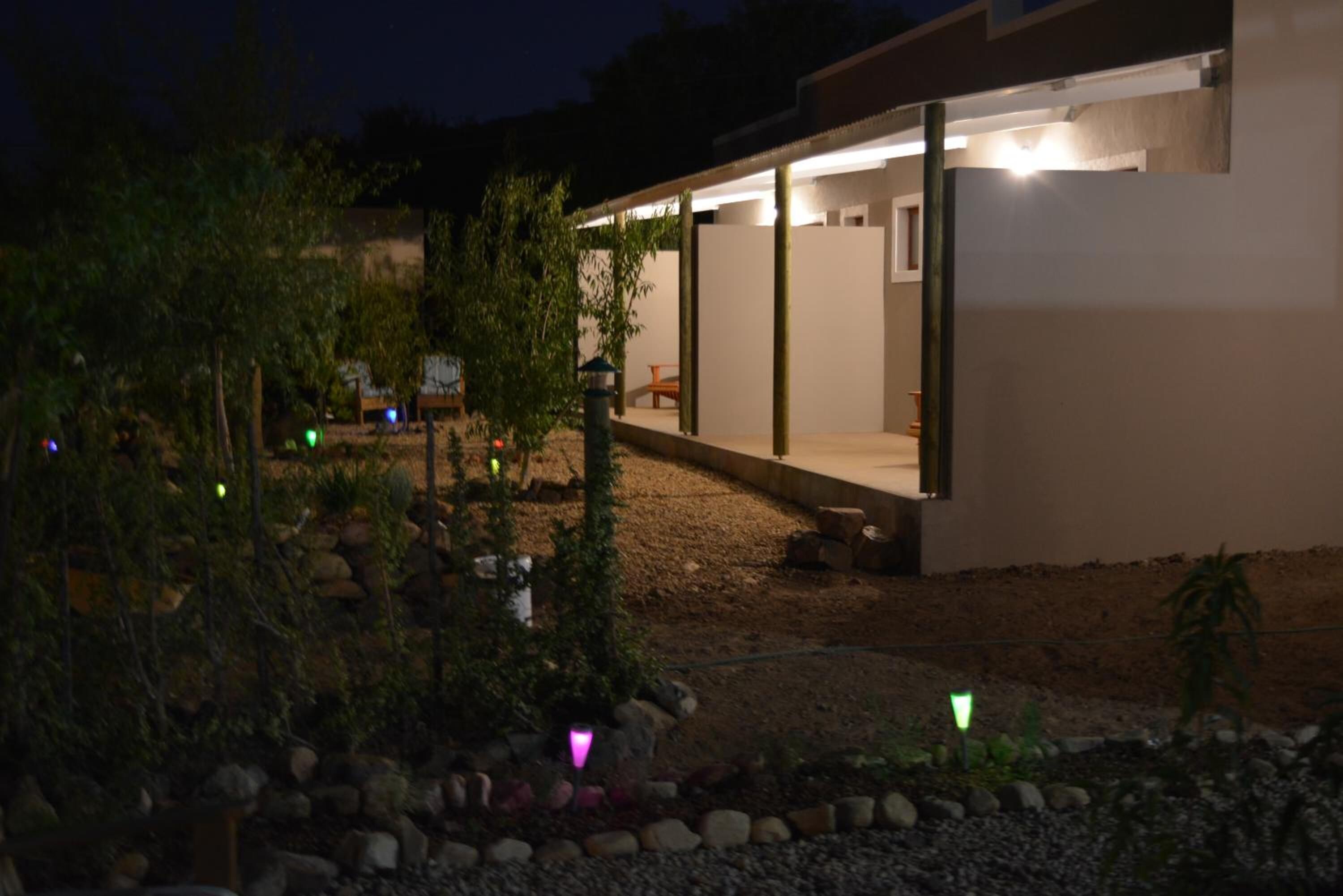 Um edifício moderno com área externa coberta, rodeado por um jardim ajardinado com diversas plantas e iluminação decorativa.