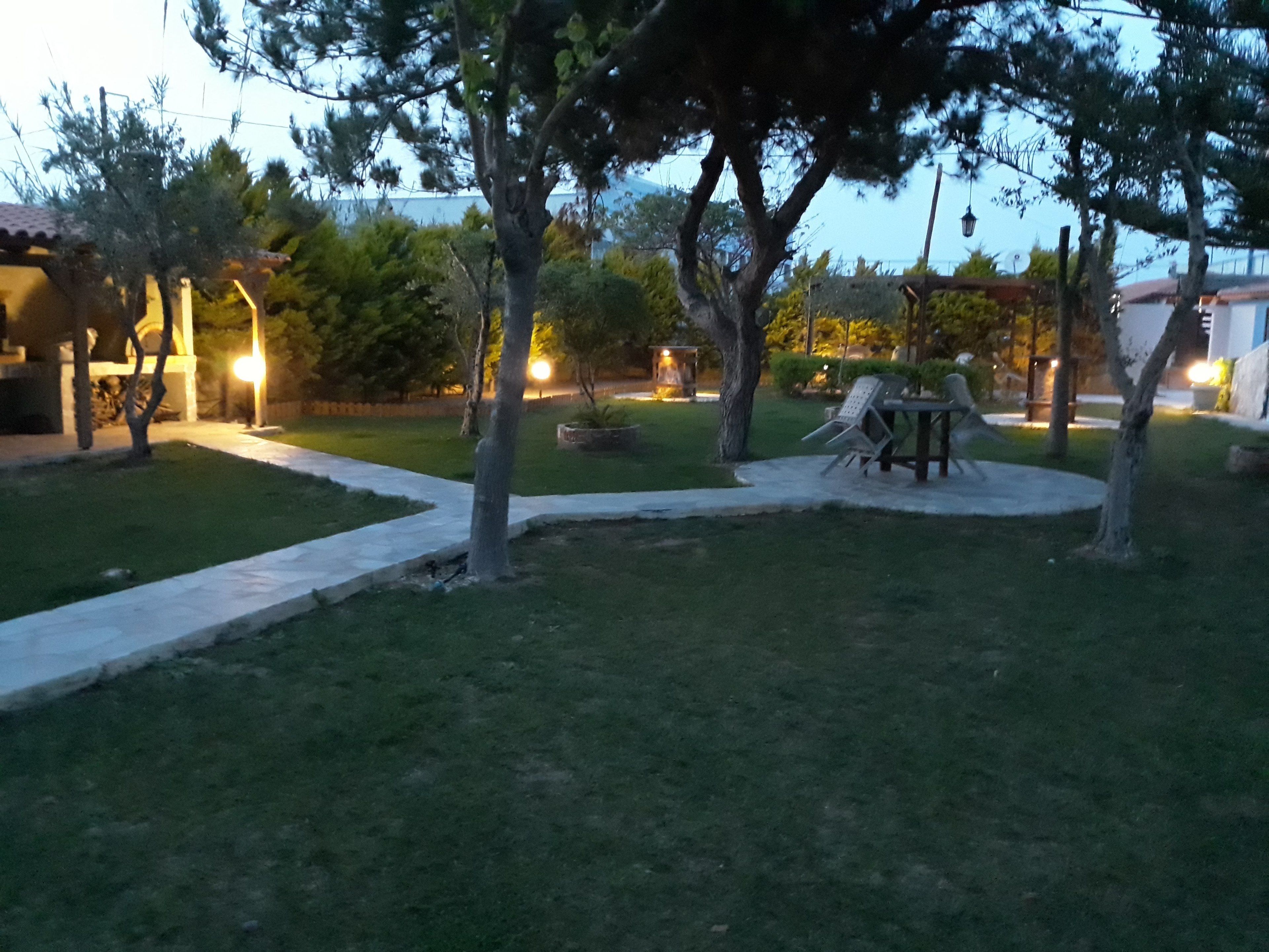 Uma área externa bem cuidada, com gramado, calçada pavimentada e espaço de estar com mesas e cadeiras.