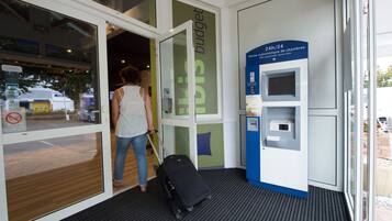 Entrada de hotel com porta de vidro, carrinho de bagagem e máquina de aluguel de salas.