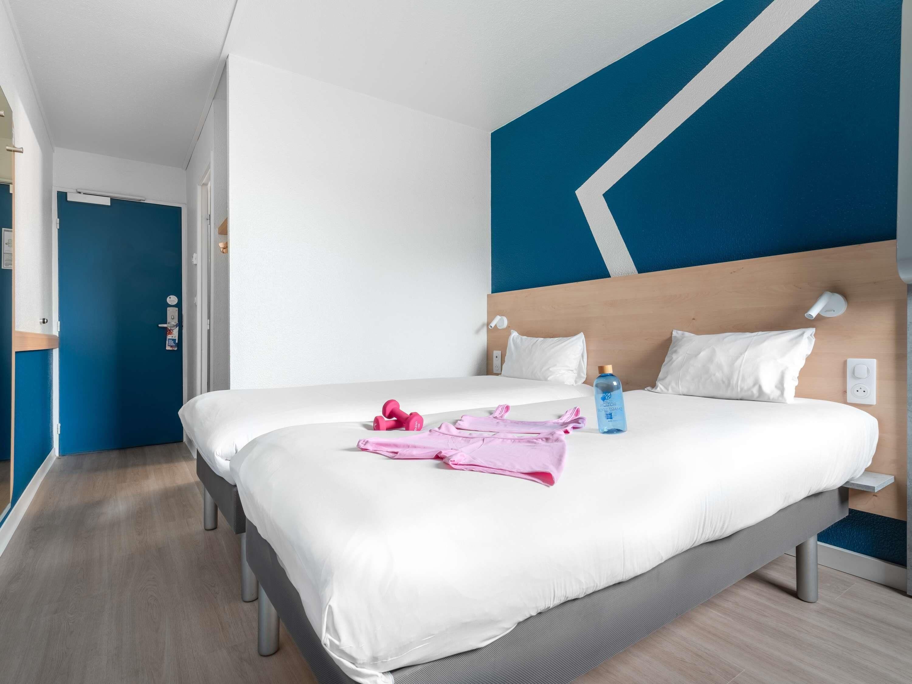 Quarto de hotel com uma cama grande, roupa de cama branca, parede azul com design geométrico e cabeceira de madeira.