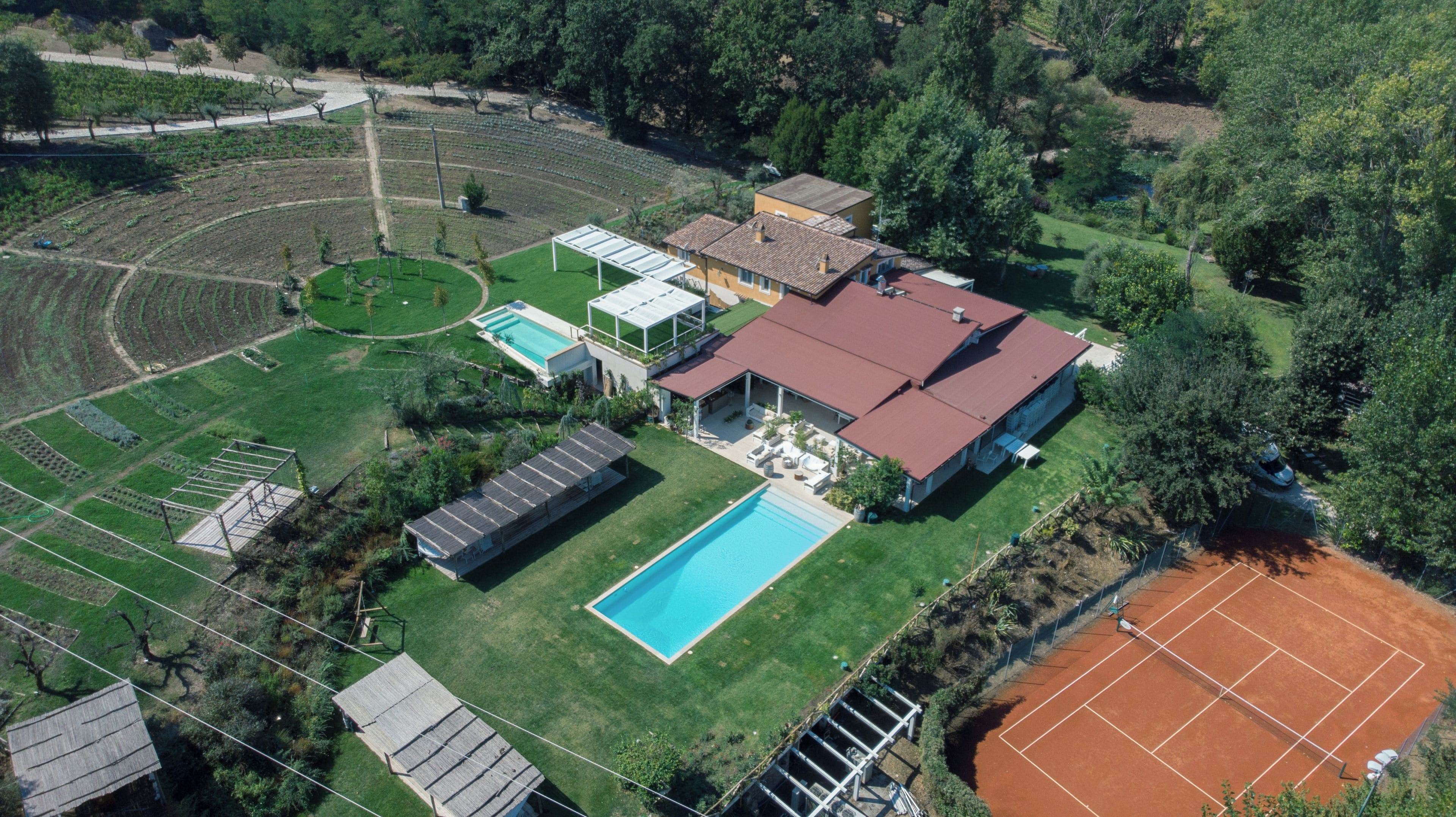 Vista aérea de uma propriedade residencial com piscina, quadra de tênis e uma casa grande com telhado vermelho.