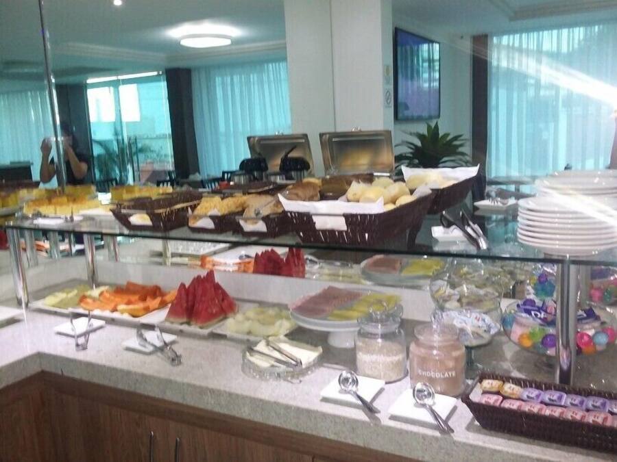 Buffet com diversas opções de comida, incluindo frutas, pastelaria e sobremesas, expostas em um balcão de vidro com utensílios de servir.