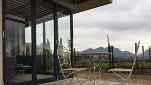 Um pátio com duas cadeiras de metal e uma mesa, uma porta de vidro que dá para o interior e vista para montanhas através de uma cerca.