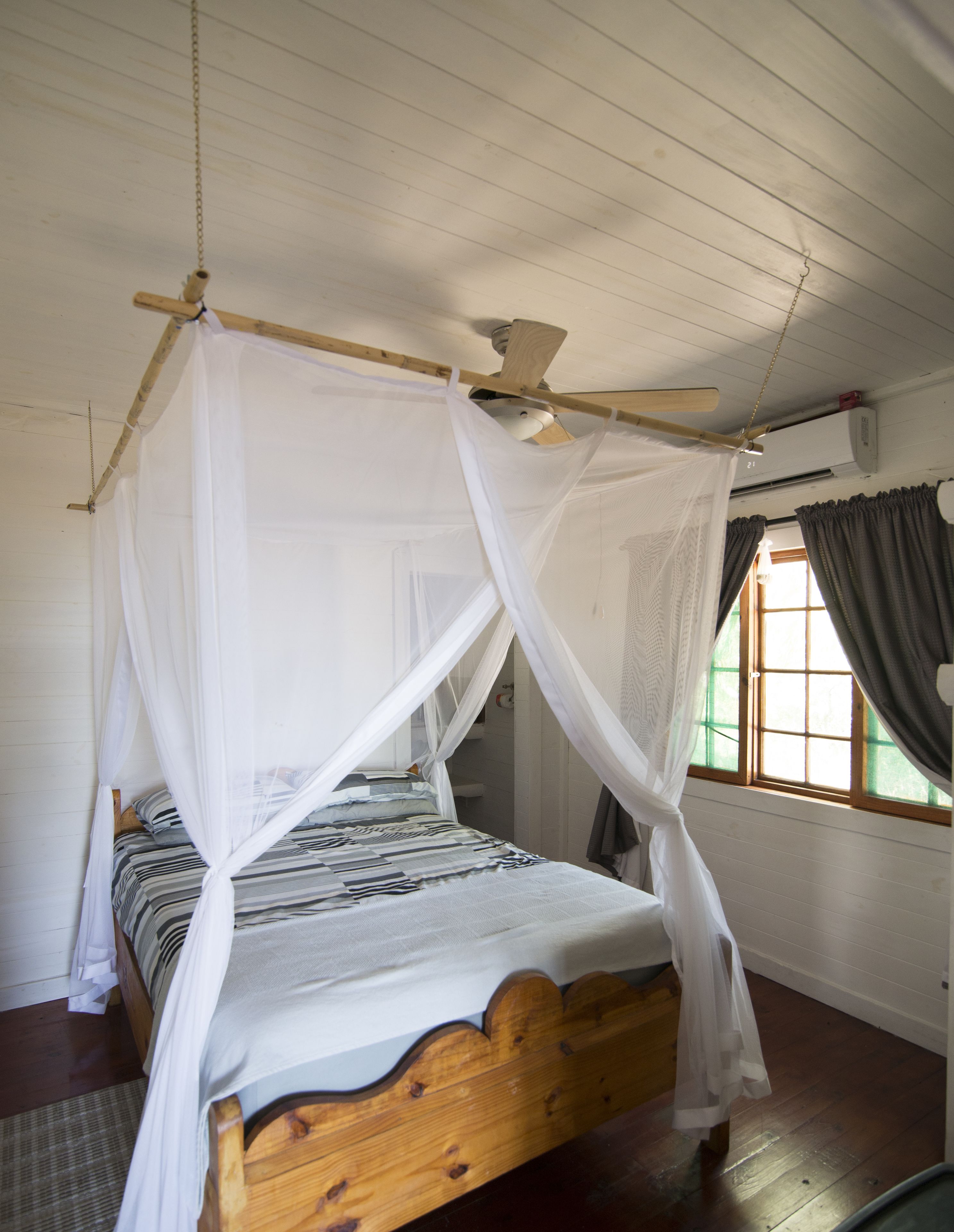 Uma cama de madeira com dossel e cortinas brancas, um ventilador de teto de madeira e uma janela com cortinas.