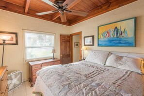 Quarto com teto de madeira, cama com colcha florida, pintura de veleiros e janela com persianas.