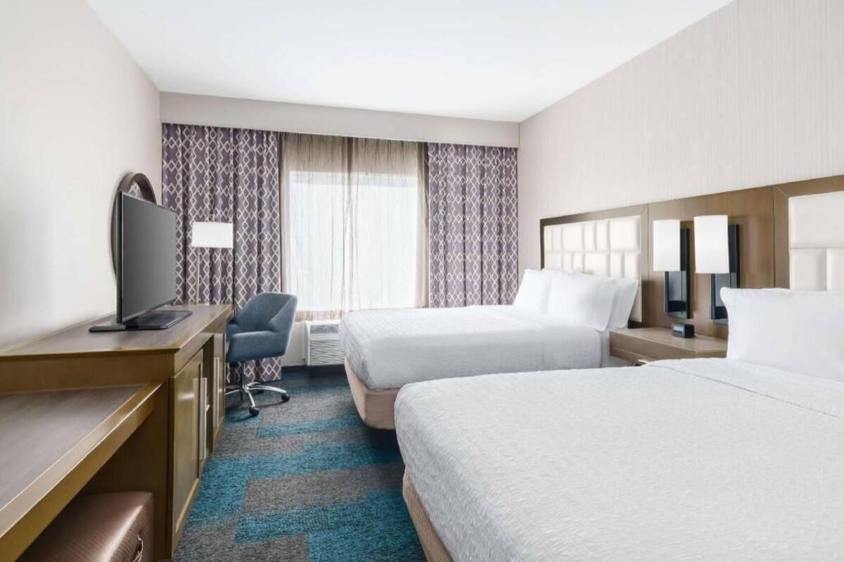 Quarto de hotel com duas camas, uma escrivaninha, uma cadeira, uma televisão e uma janela com cortinas.