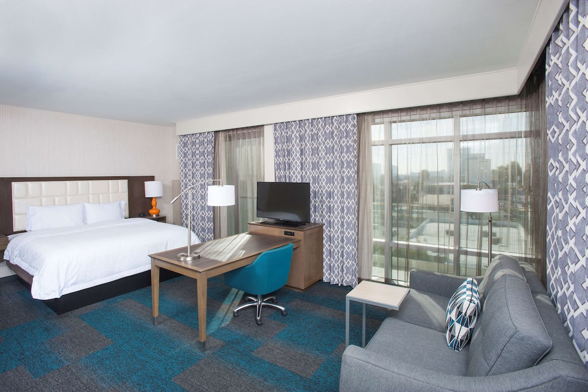 Quarto de hotel com uma cama, uma escrivaninha com cadeira, televisão e um sofá.