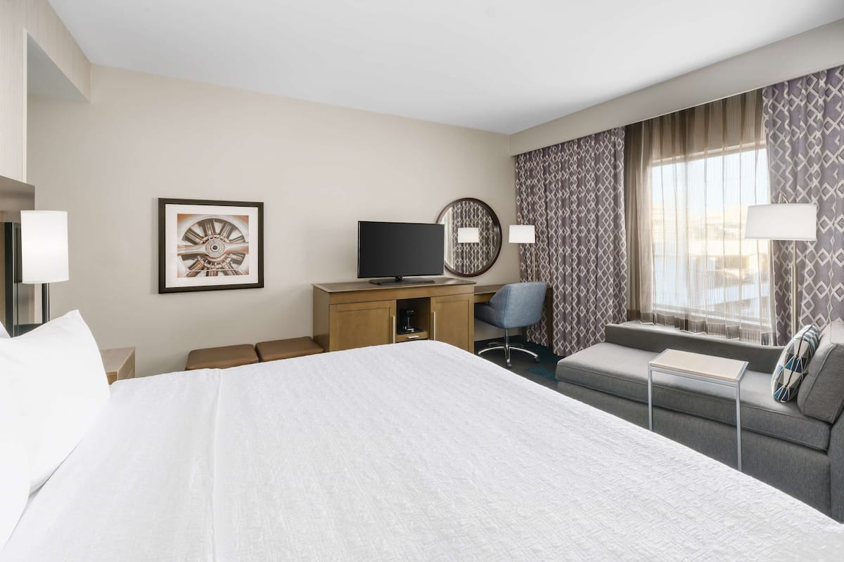 Quarto de hotel com uma cama grande, uma escrivaninha com televisão, uma poltrona, um sofá e uma janela com cortinas.