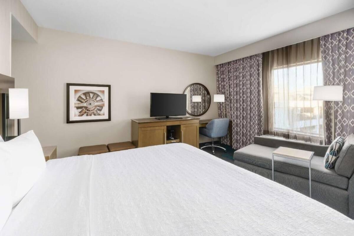 Quarto de hotel com uma cama grande, uma escrivaninha com televisão, uma cadeira, um sofá e uma janela com cortinas.