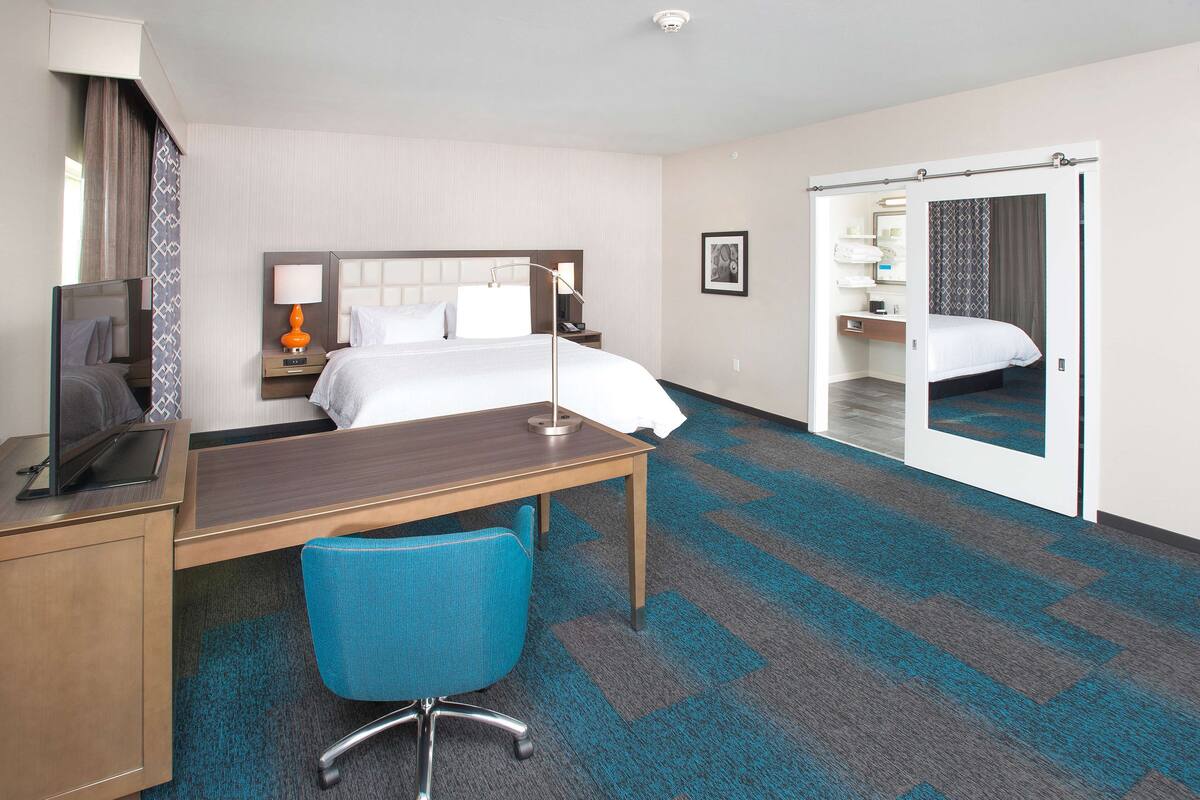 Quarto de hotel com cama, escrivaninha, cadeira e espelho.
