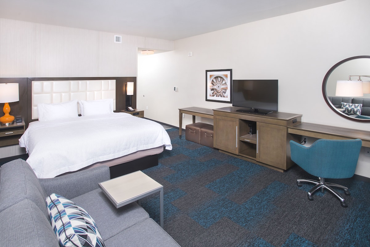 Quarto de hotel com cama, escrivaninha, cadeira, sofá, televisão e espelho.