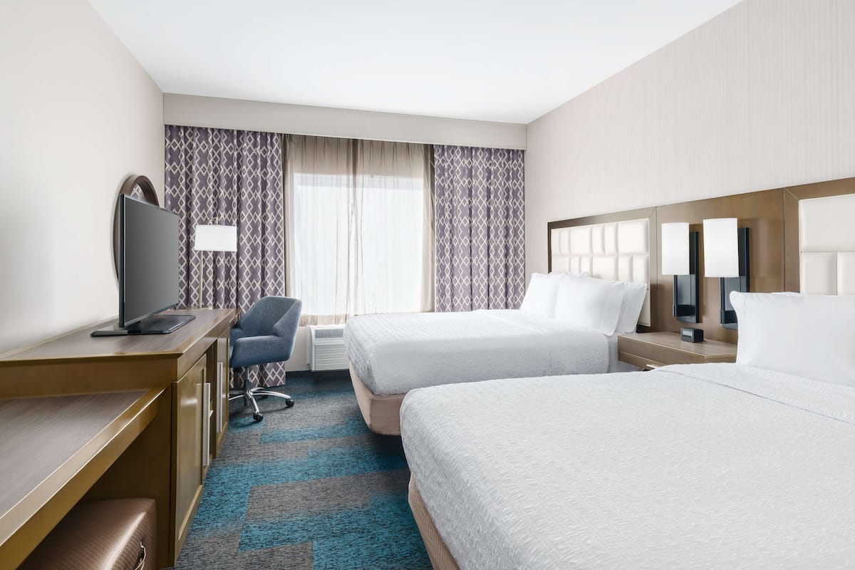 Quarto de hotel com duas camas, uma escrivaninha, uma cadeira, uma televisão e uma janela com cortinas.