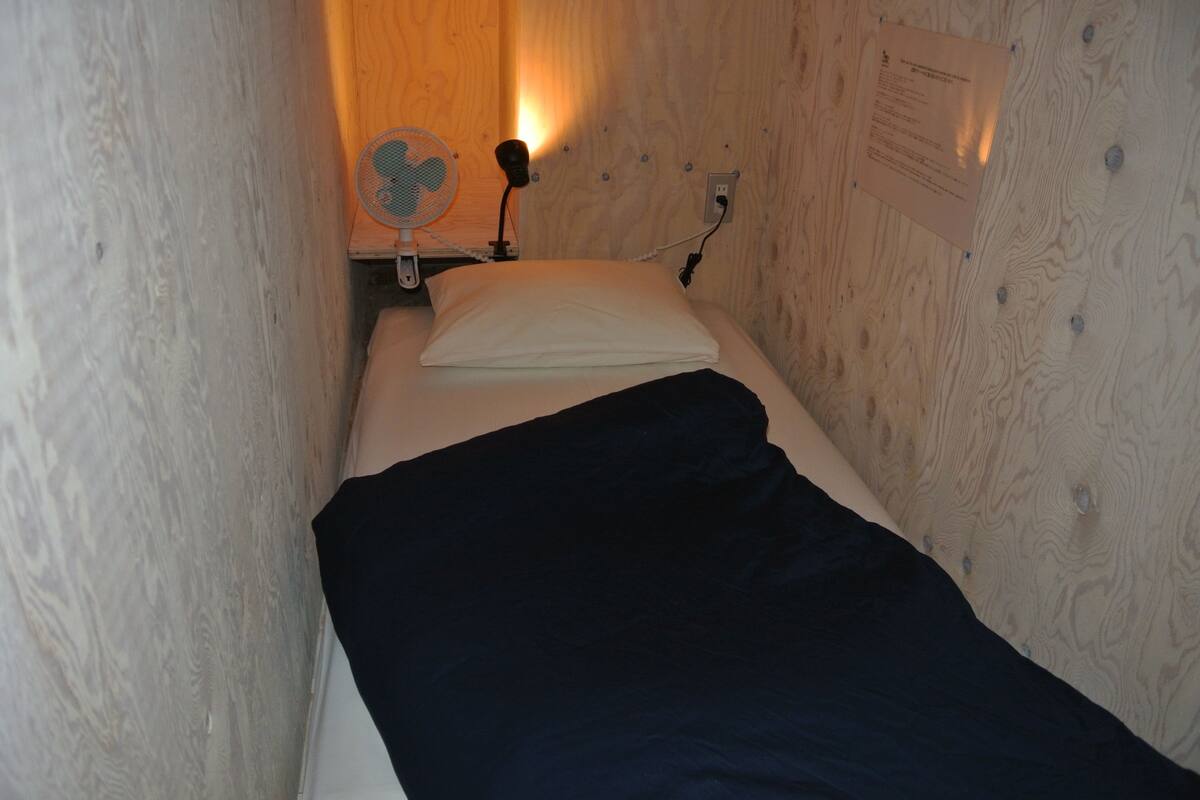 Quarto estreito com painéis de madeira, uma cama simples, um ventilador na parede e uma pequena mesa com abajur.